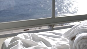 Cama com lençóis brancos e vista para o oceano através de uma janela.