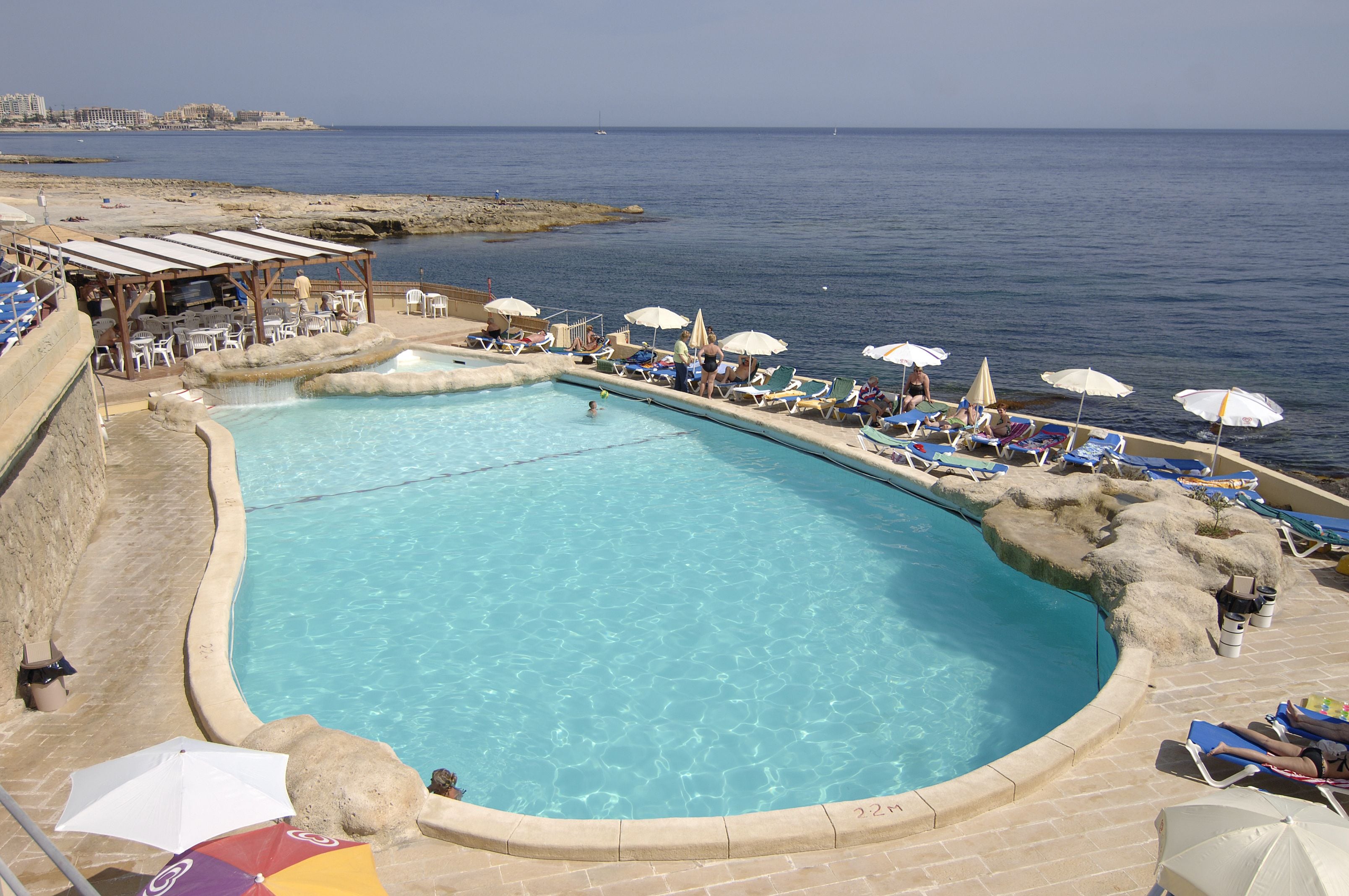 Piscina com água azul cristalina, rodeada por espreguiçadeiras e guarda-sóis, com vista para o mar e uma costa rochosa.