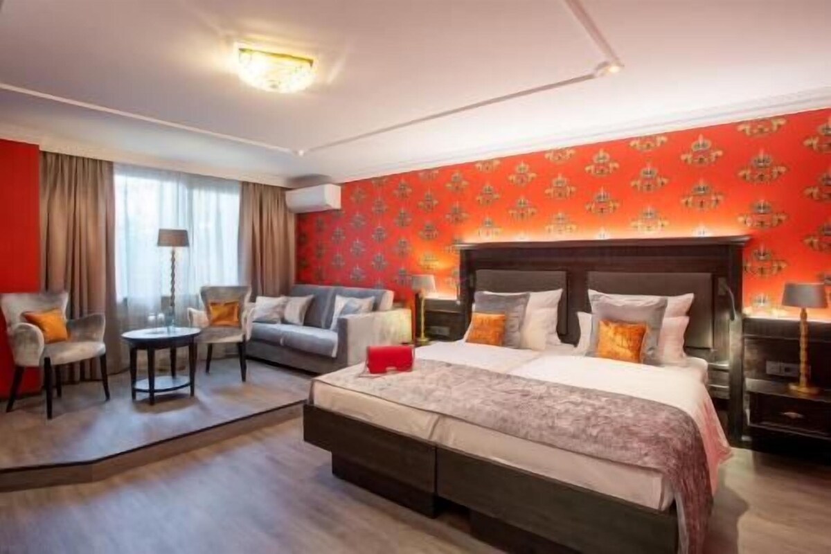 Quarto de hotel com uma cama grande, um sofá, uma cadeira, uma mesinha e uma parede de destaque vermelha.
