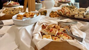 Mesa de buffet com diversos alimentos, uma planta em vaso e vista para o verde lá fora.