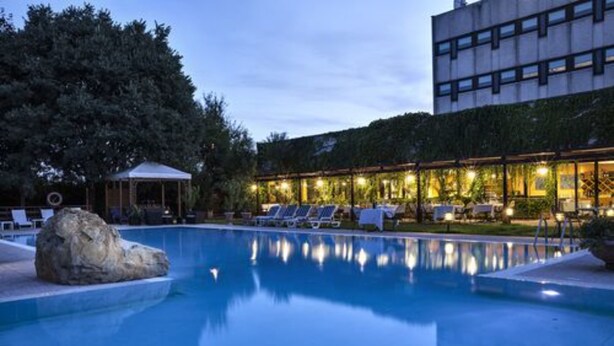Uma piscina com espreguiçadeiras, um elemento decorativo de rochas e um edifício com janelas iluminadas ao fundo.