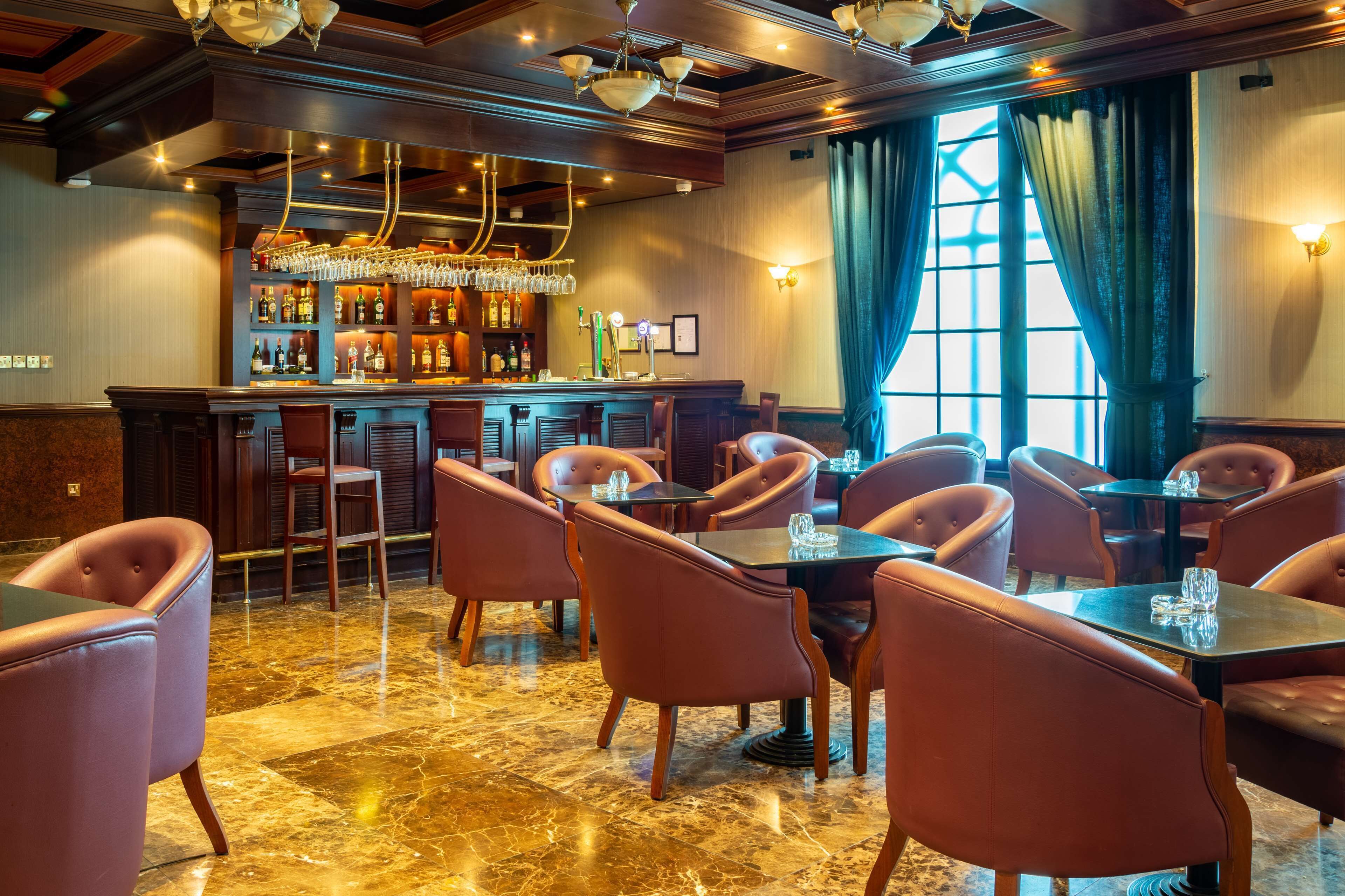 Uma área de bar com piso de mármore, cadeiras de couro e balcão com garrafas e copos.