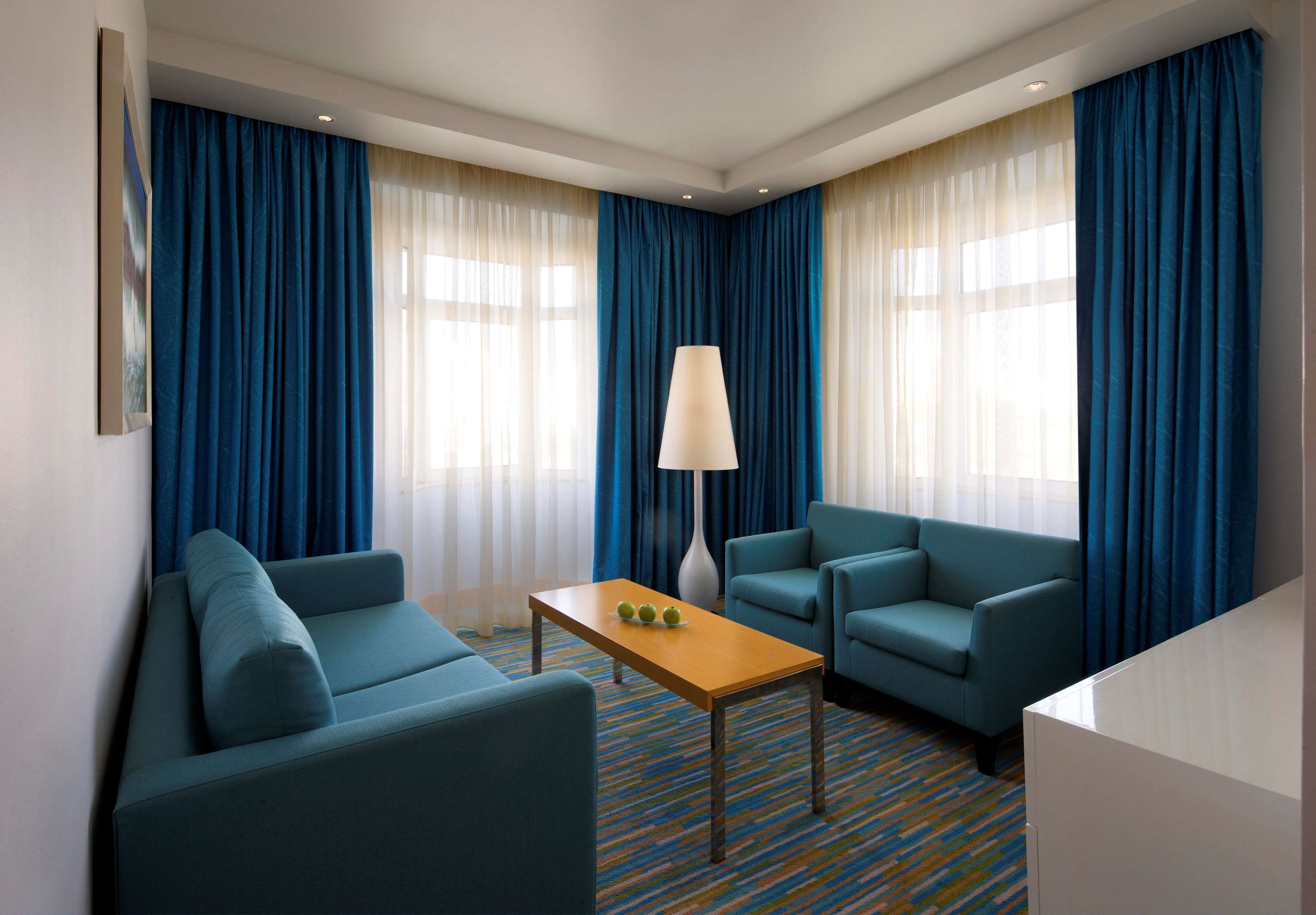 Quarto de hotel com sofá, duas poltronas, uma mesa com maçãs, abajur e vista através de cortinas finas.
