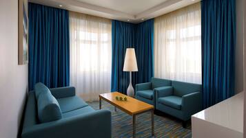Quarto de hotel com sofá, duas poltronas, uma mesa com maçãs, abajur e vista através de cortinas finas.