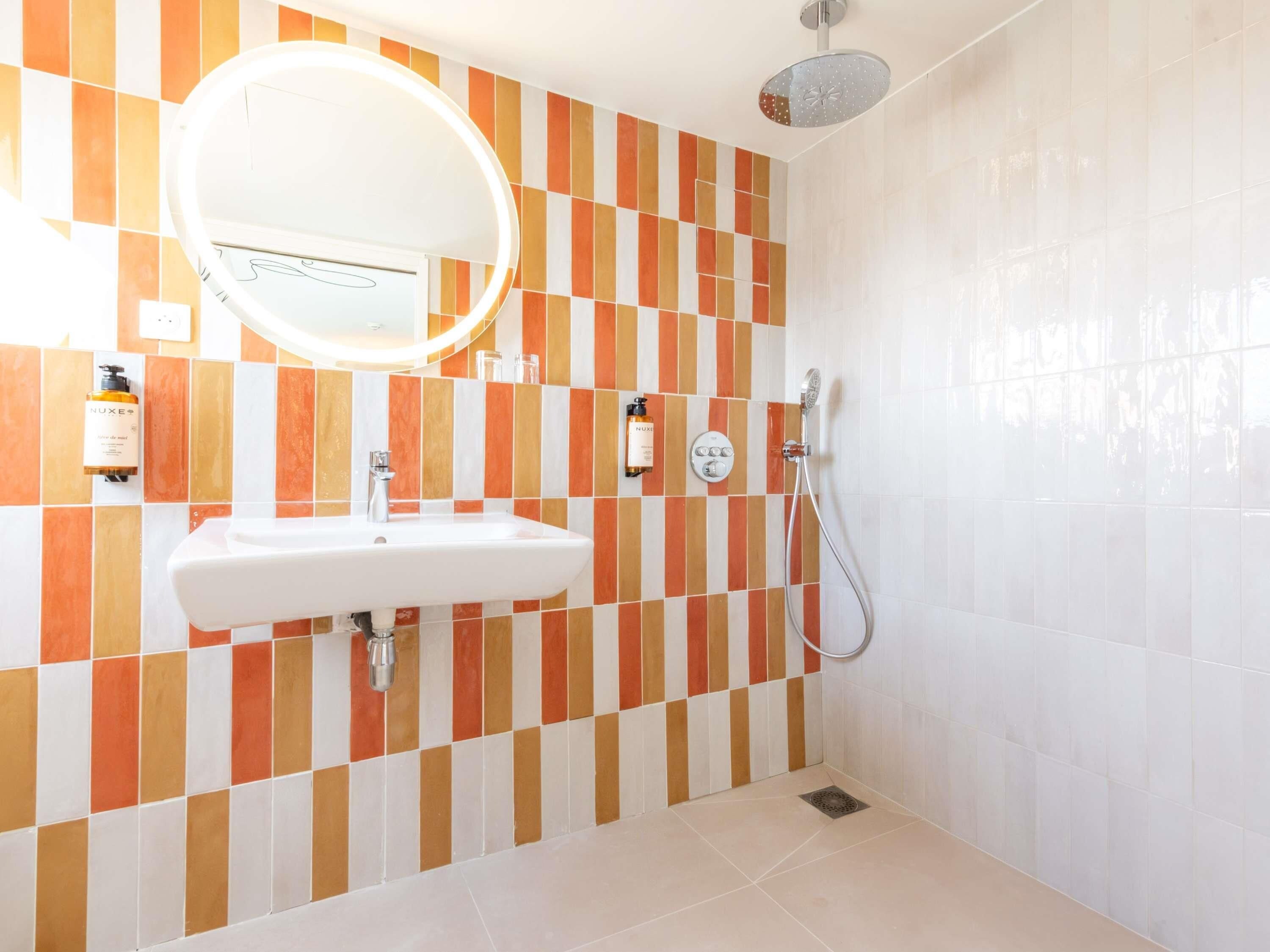 Casa de banho com lavatório branco, espelho redondo, chuveiro e parede revestida a mosaicos xadrez.