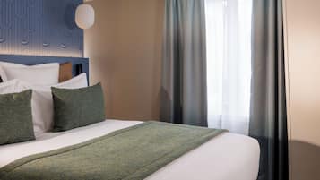 Quarto de hotel com cama grande, cabeceira azul, janela com cortinas e piso coberto por carpete.