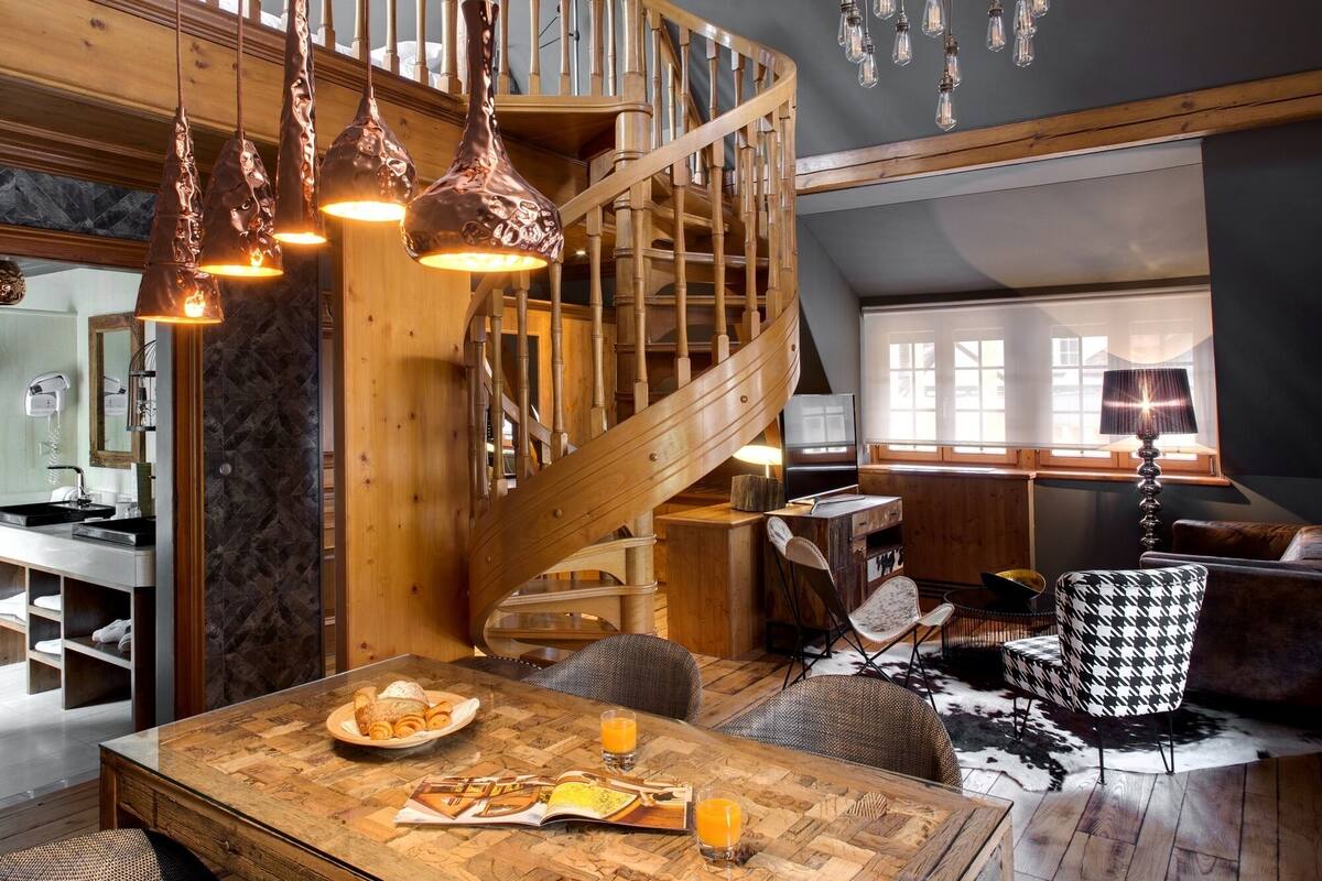 Uma ampla sala de estar com escada de madeira, mesa de jantar com comida e bebidas, e um espaço de estar acolhedor com almofadas estampadas.