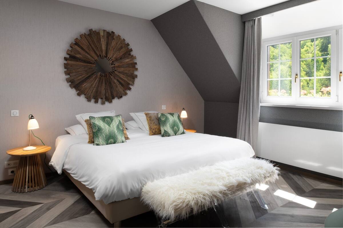 Um quarto moderno com uma cama grande, duas mesas de cabeceira, um detalhe decorativo na parede e uma janela com cortinas.