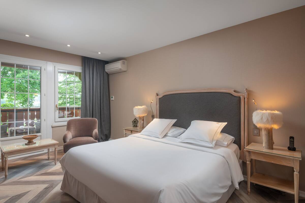 Quarto de hotel com uma cama grande, duas mesas de cabeceira com abajures, uma cadeira, uma pequena mesa com uma tigela e uma janela com cortinas.