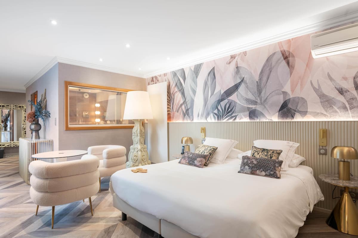 Quarto de hotel moderno com uma cama grande, uma mesa redonda, uma cadeira e um espelho decorativo.