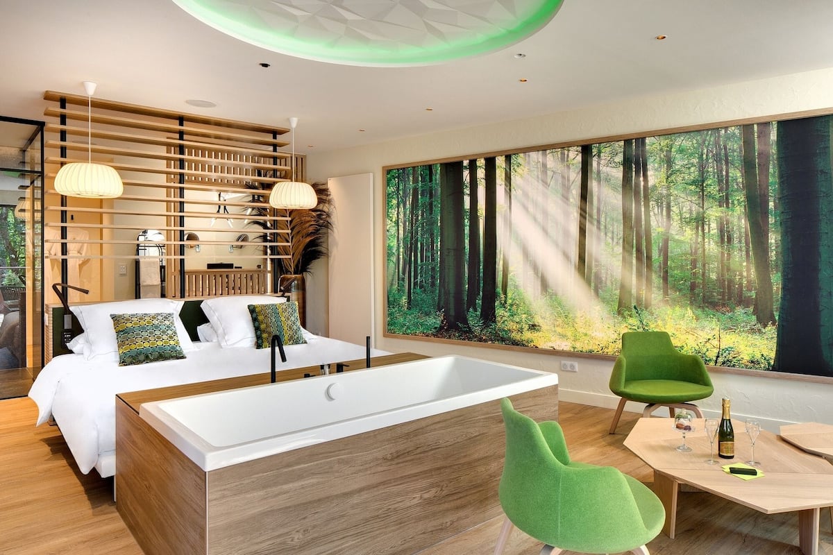 Quarto moderno com uma cama grande, uma banheira e uma janela ampla com vista para uma floresta.