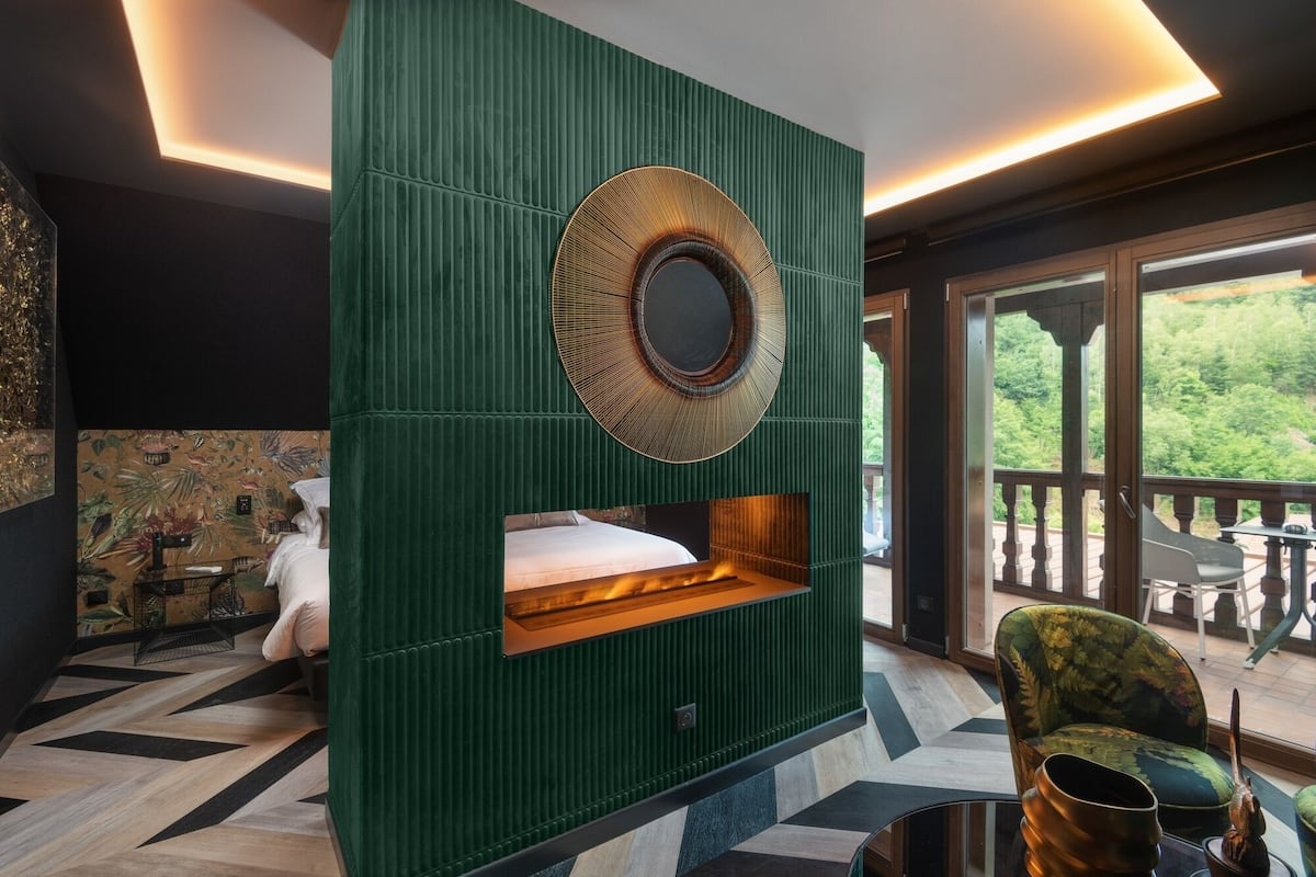Quarto com parede verde, espelho circular, cama e vista para o exterior com vegetação.