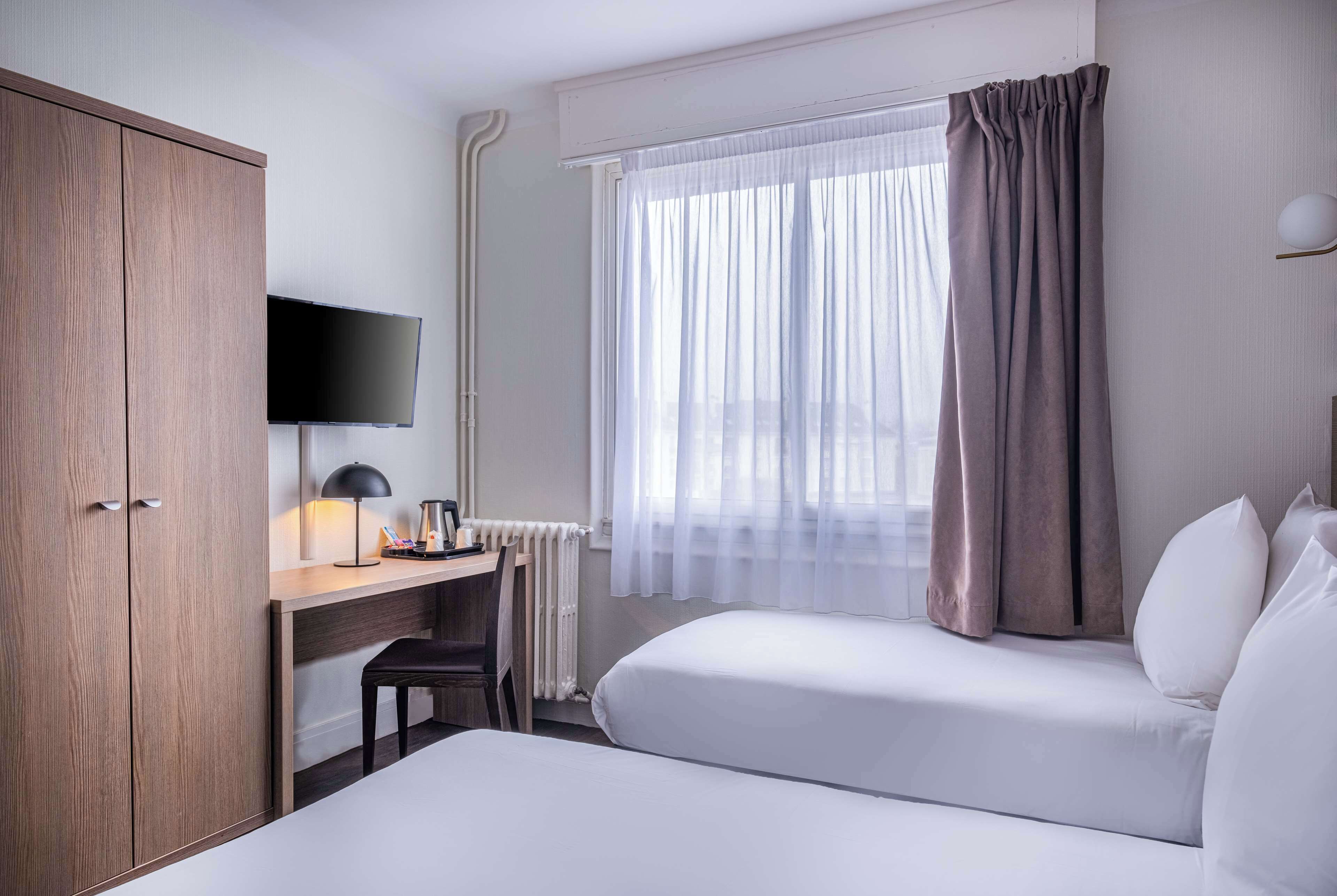 Quarto de hotel com guarda-roupa de madeira, secretária com computador e luminária, duas camas, janela com cortinas leves e radiador.