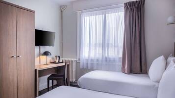 Quarto de hotel com guarda-roupa de madeira, secretária com computador e luminária, duas camas, janela com cortinas leves e radiador.