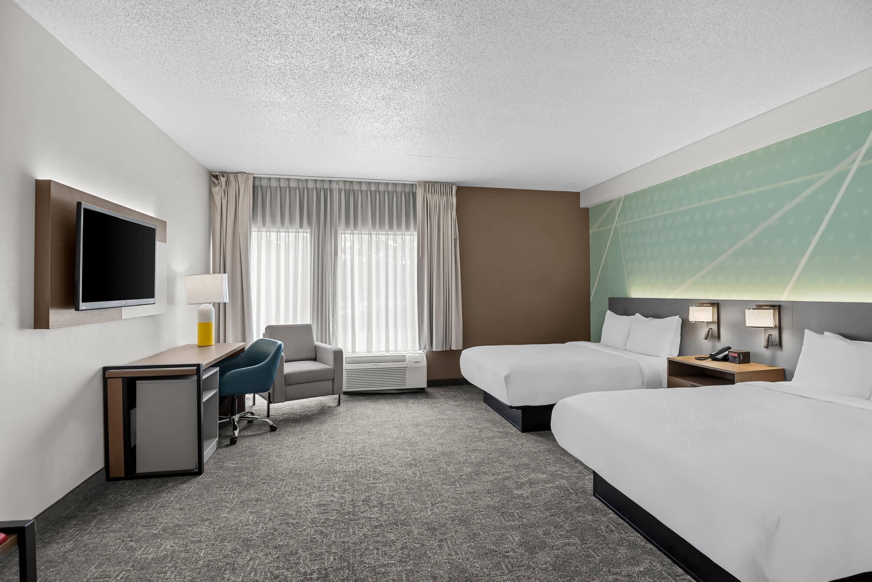 Quarto de hotel com duas camas, uma escrivaninha com cadeira, televisão e janela com cortinas.