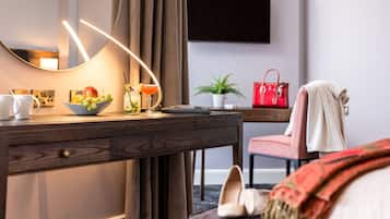 Quarto de hotel com uma escrivaninha de madeira, um espelho, uma cesta de fruta, uma bolsa vermelha, uma planta e uma televisão na parede.