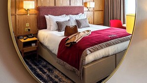 Quarto de hotel com um espelho redondo grande refletindo a cama, painéis de madeira, uma cadeira vermelha e uma mesa de cabeceira com abajur e telefone.