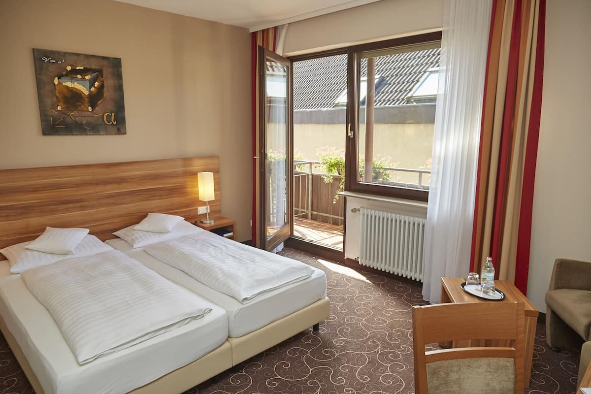 Quarto de hotel com duas camas, cabeceira de madeira, uma mesinha com uma garrafa e copos, e vista para a varanda externa.