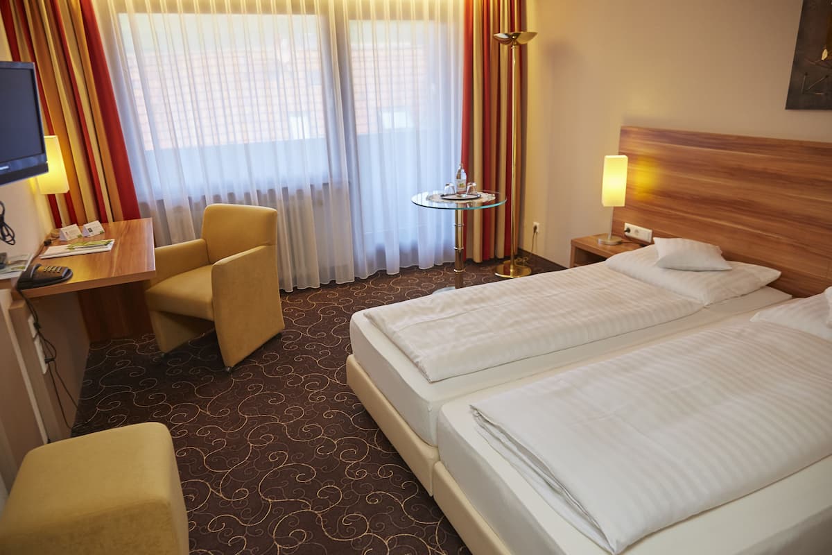 Quarto de hotel com duas camas, uma escrivaninha, uma cadeira, uma mesinha e uma televisão.