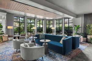 Um hall amplo com um sofá azul, poltronas cinzas e mesas redondas, janelas grandes com vista para o verde.
