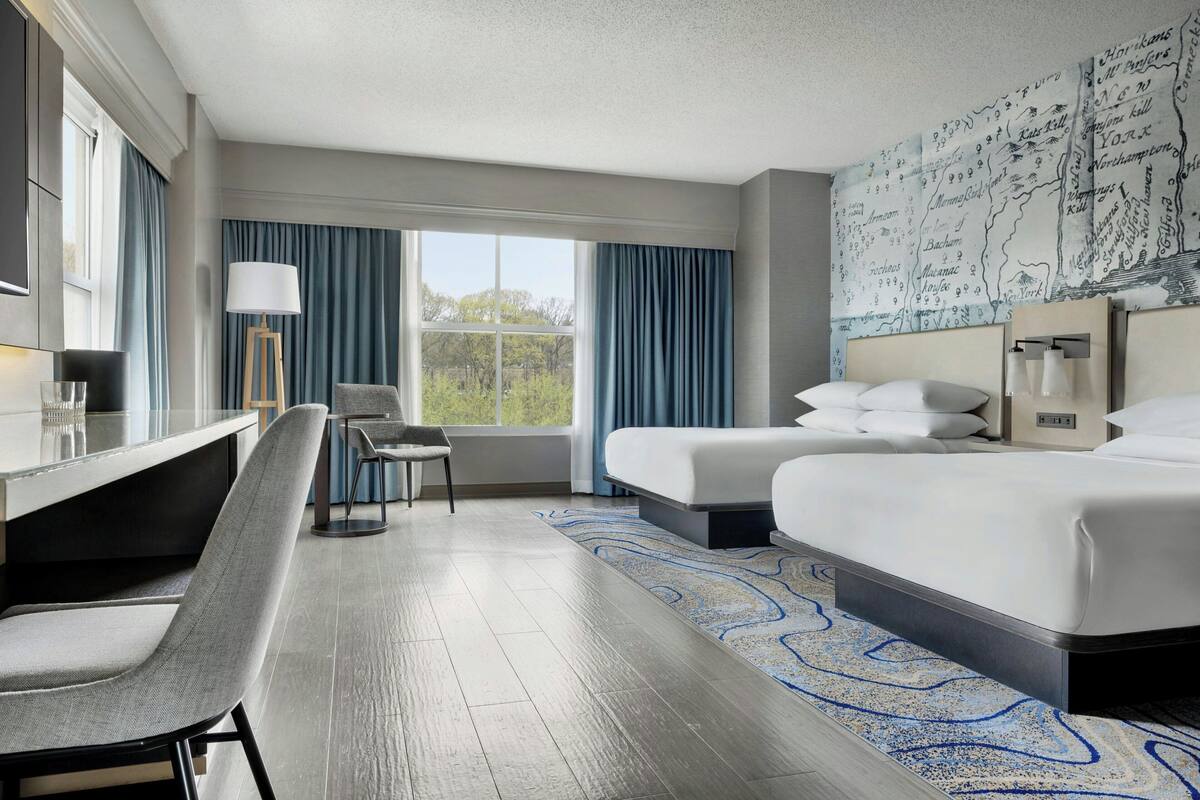 Quarto de hotel com escrivaninha, duas camas, uma cadeira, abajur e uma janela grande com cortinas.