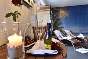Uma mesa com vela, garrafa de champanhe e copos, com um mural ao fundo retratando uma cena de praia.