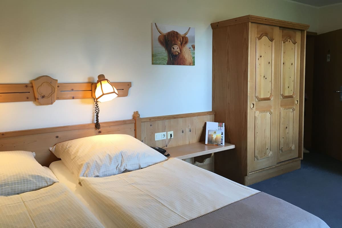 Quarto de hotel com duas camas, cabeceira de madeira, luminária fixada na parede, guarda-roupa de madeira e um quadro com uma vaca.