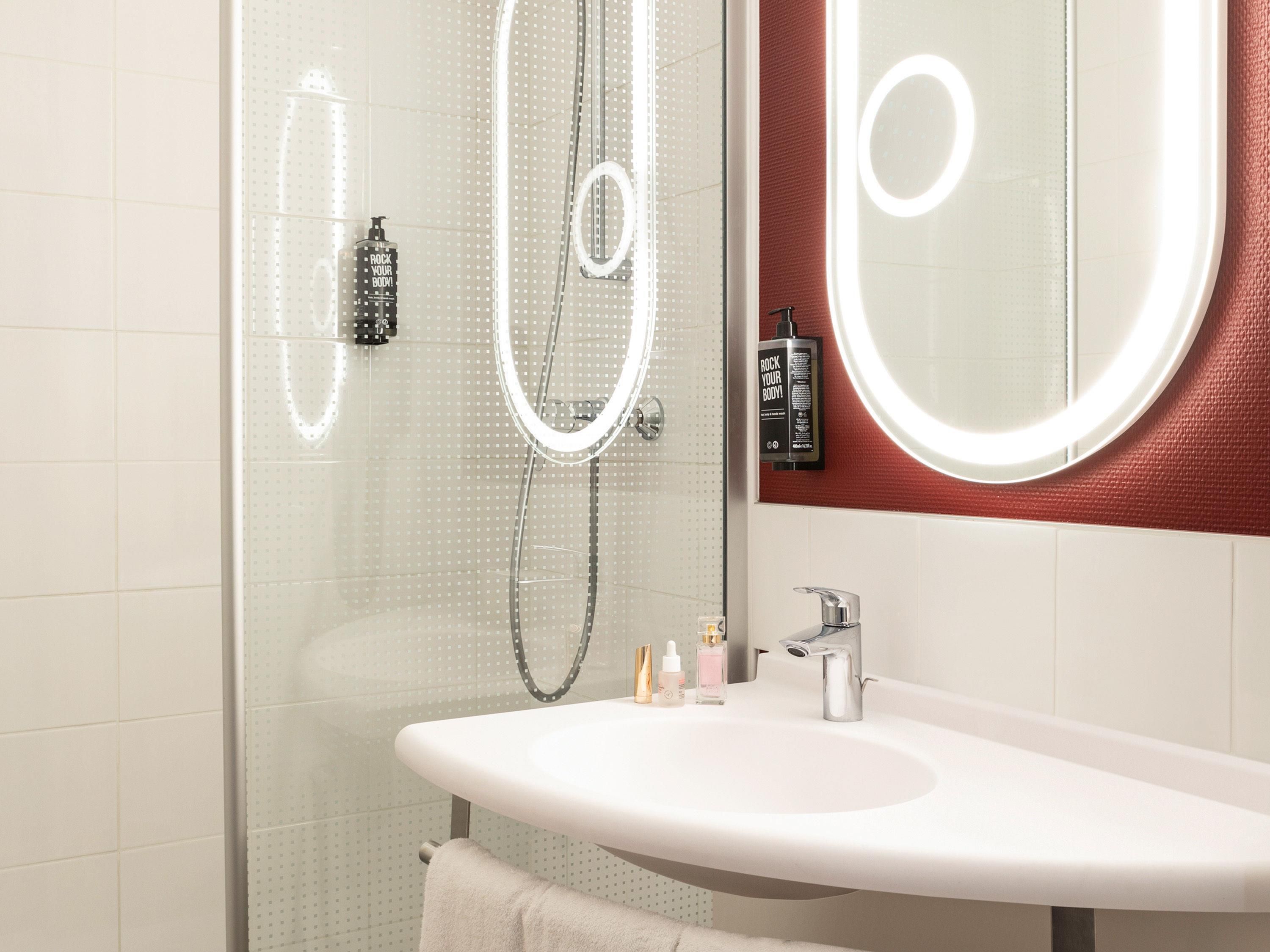 Uma casa de banho com cabine de duche em vidro, lavatório branco com torneira moderna e espelho com iluminação circular.