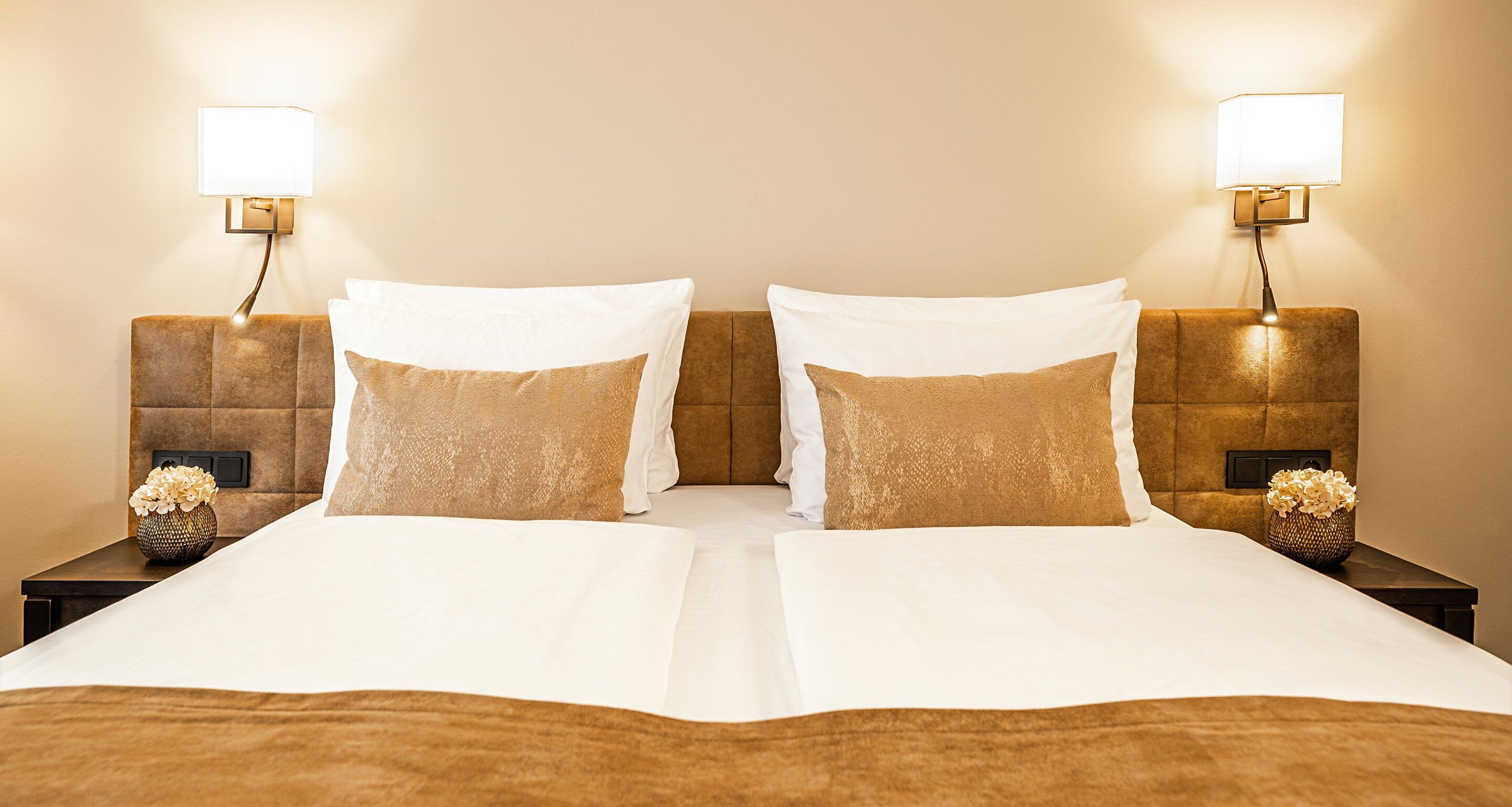 Uma cama de casal com roupa de cama branca e castanha, duas mesas de cabeceira com luminárias e vasos decorativos em cada lado.