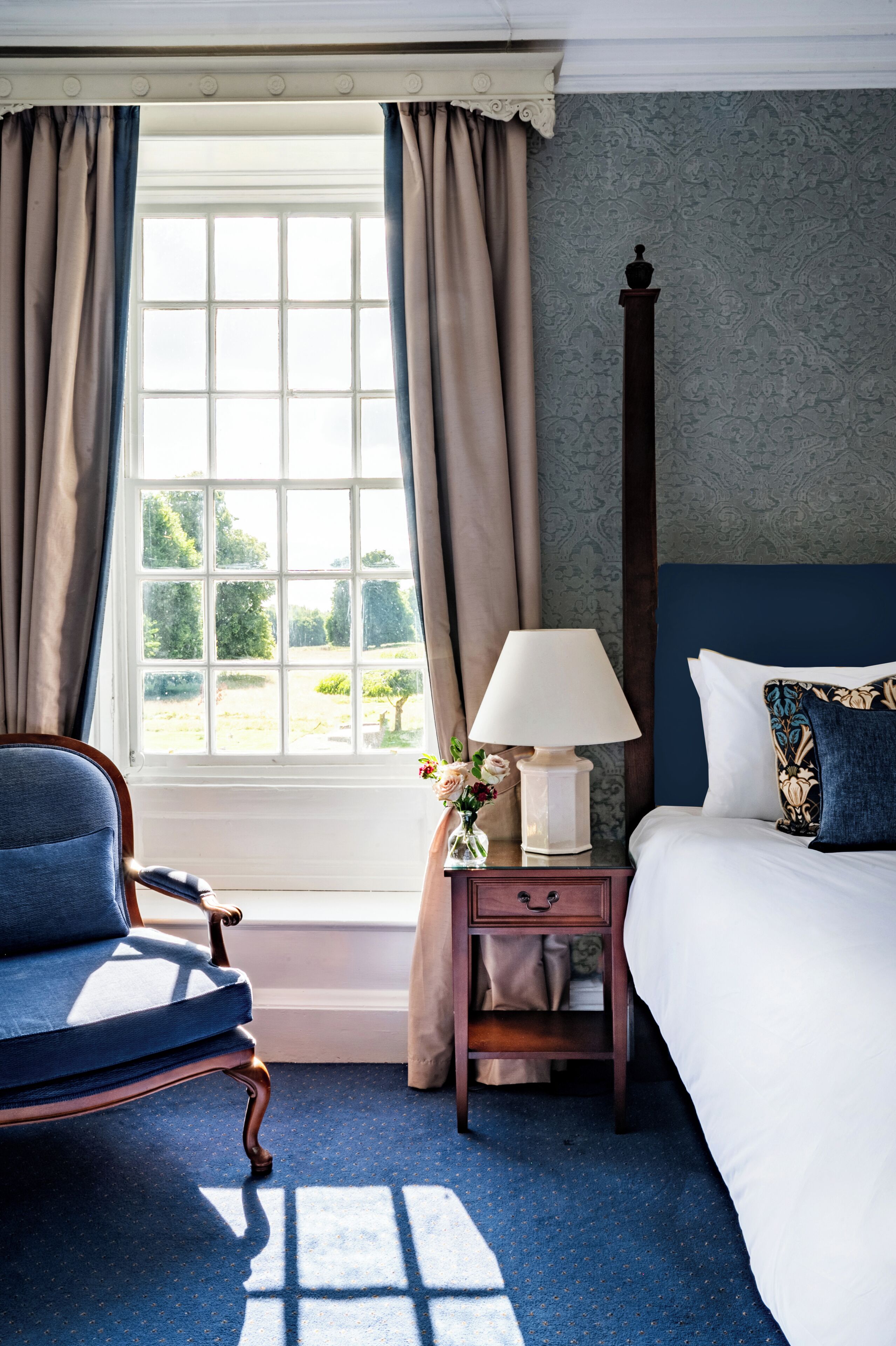 Quarto de hotel com cama, mesa de cabeceira com abajur, cadeira e janela com cortinas.
