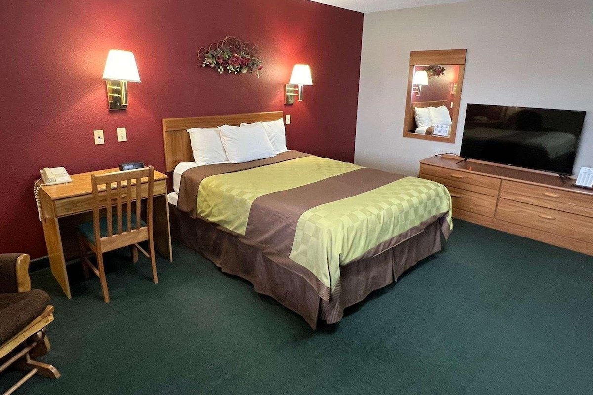 Quarto de hotel com cama, abajures, escrivaninha com cadeira, televisão e cômoda.