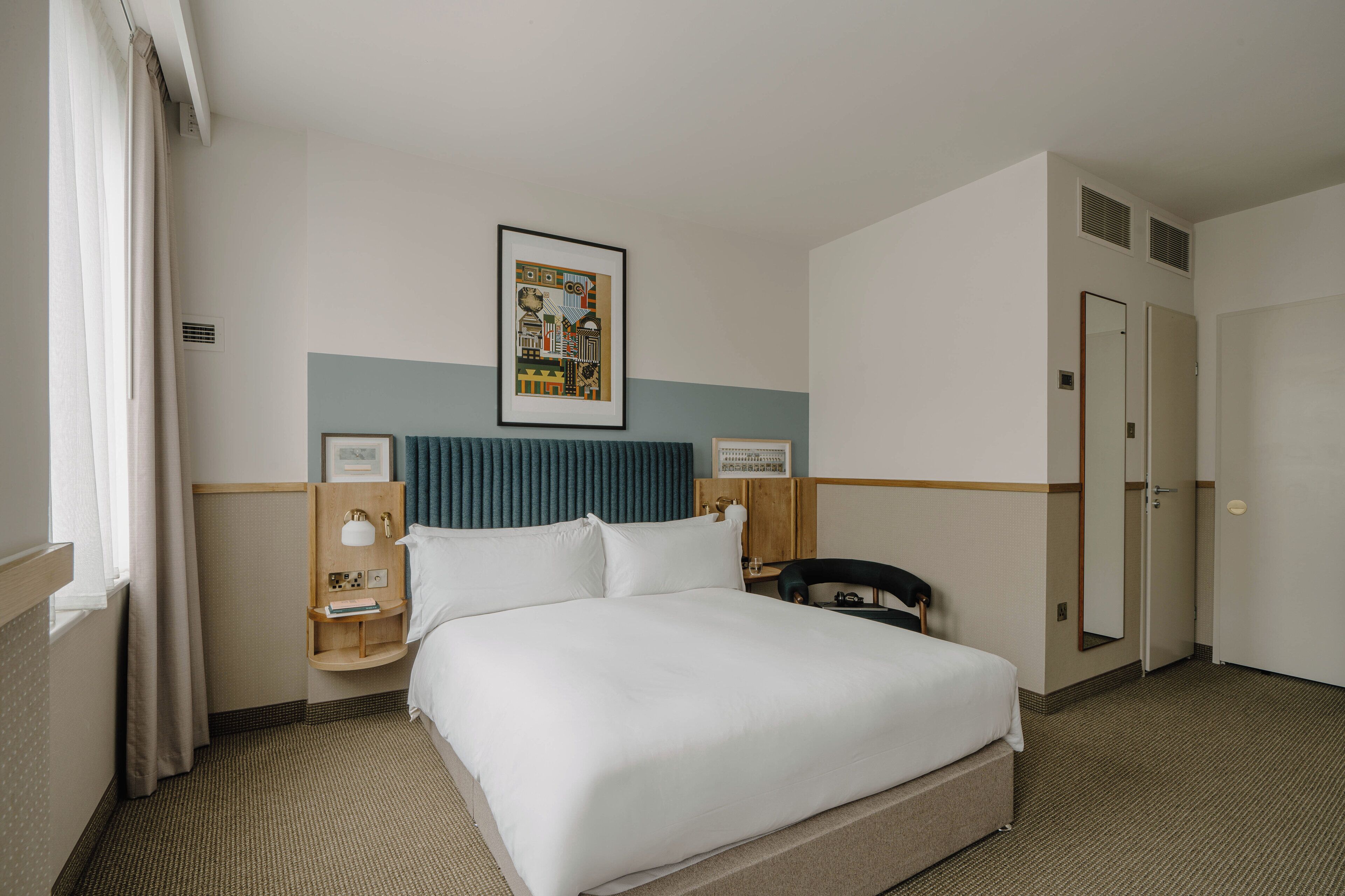 Quarto de hotel com uma cama grande, cabeceira com painel azul, quadro na parede, mesa de cabeceira com abajur e uma porta à direita.
