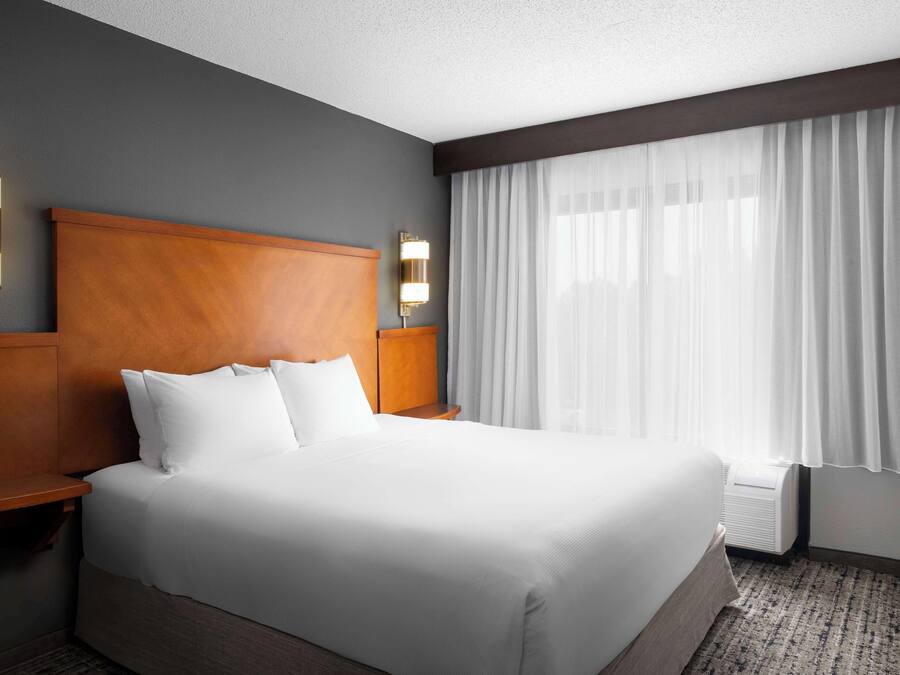 Quarto de hotel com uma cama grande, cabeceira de madeira, duas mesas de cabeceira com luminárias, uma janela ampla com cortinas translúcidas e piso carpeteado.