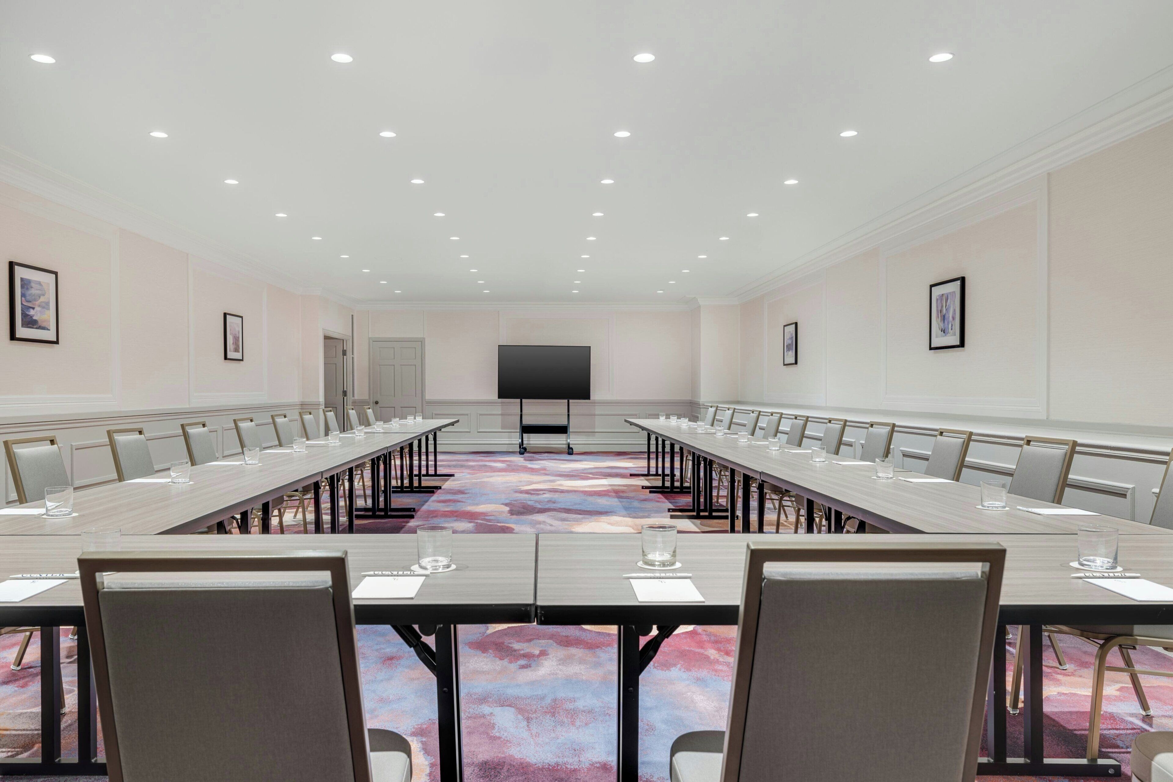 Uma sala de conferências com mesas compridas, cadeiras, um grande ecrã e quadros nas paredes.