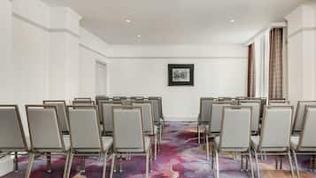 Uma sala de conferência com fileiras de cadeiras, um tapete colorido, paredes brancas e um quadro emoldurado na parede.