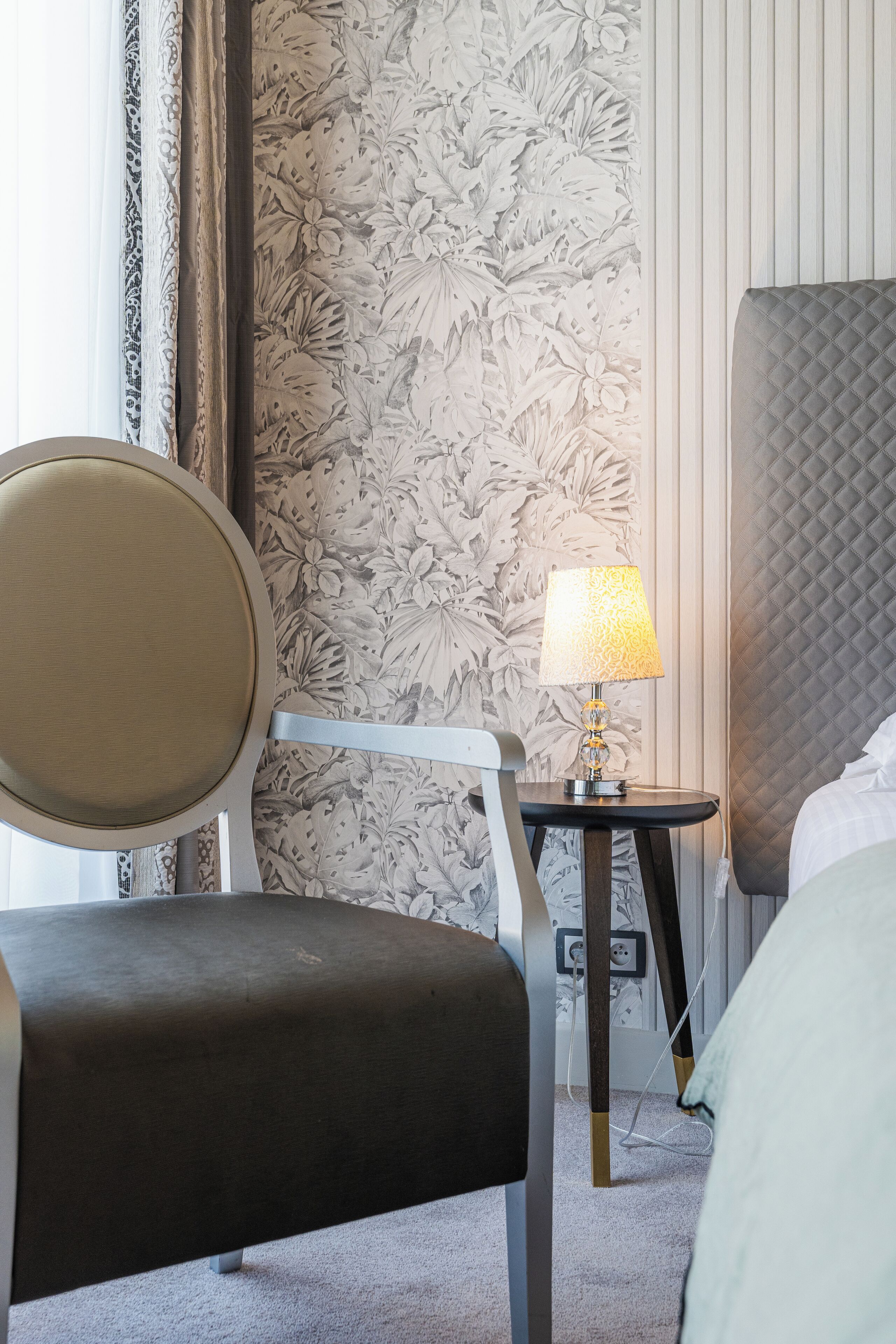 Quarto de hotel com cama, uma cadeira, uma mesa com abajur e papel de parede floral.