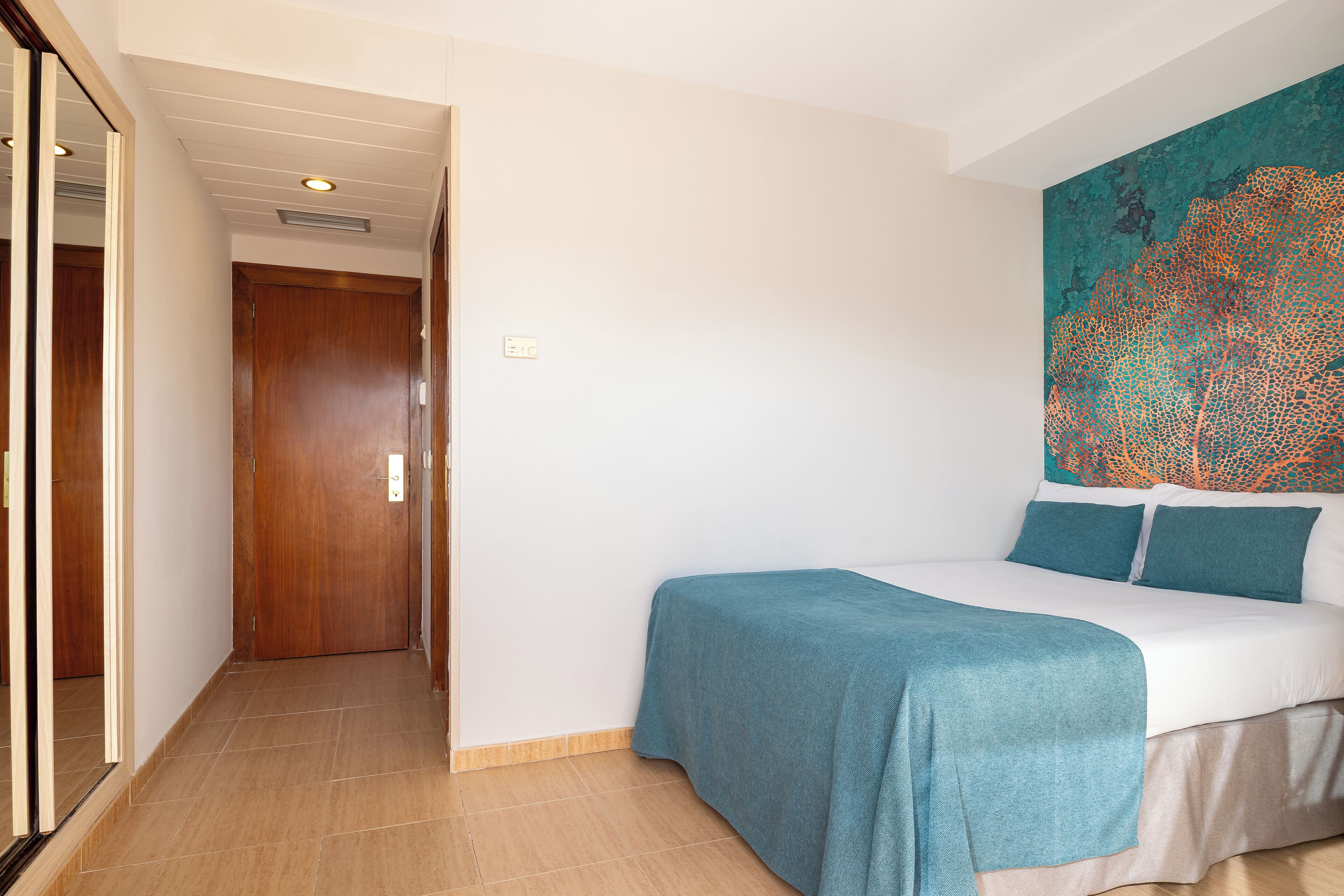 Quarto de hotel com uma cama, um cobreleito verde-água, um quadro abstrato grande na parede e uma porta de madeira.