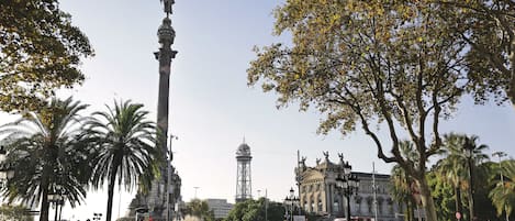 Uma praça com um monumento alto, palmeiras e uma torre ao longe.