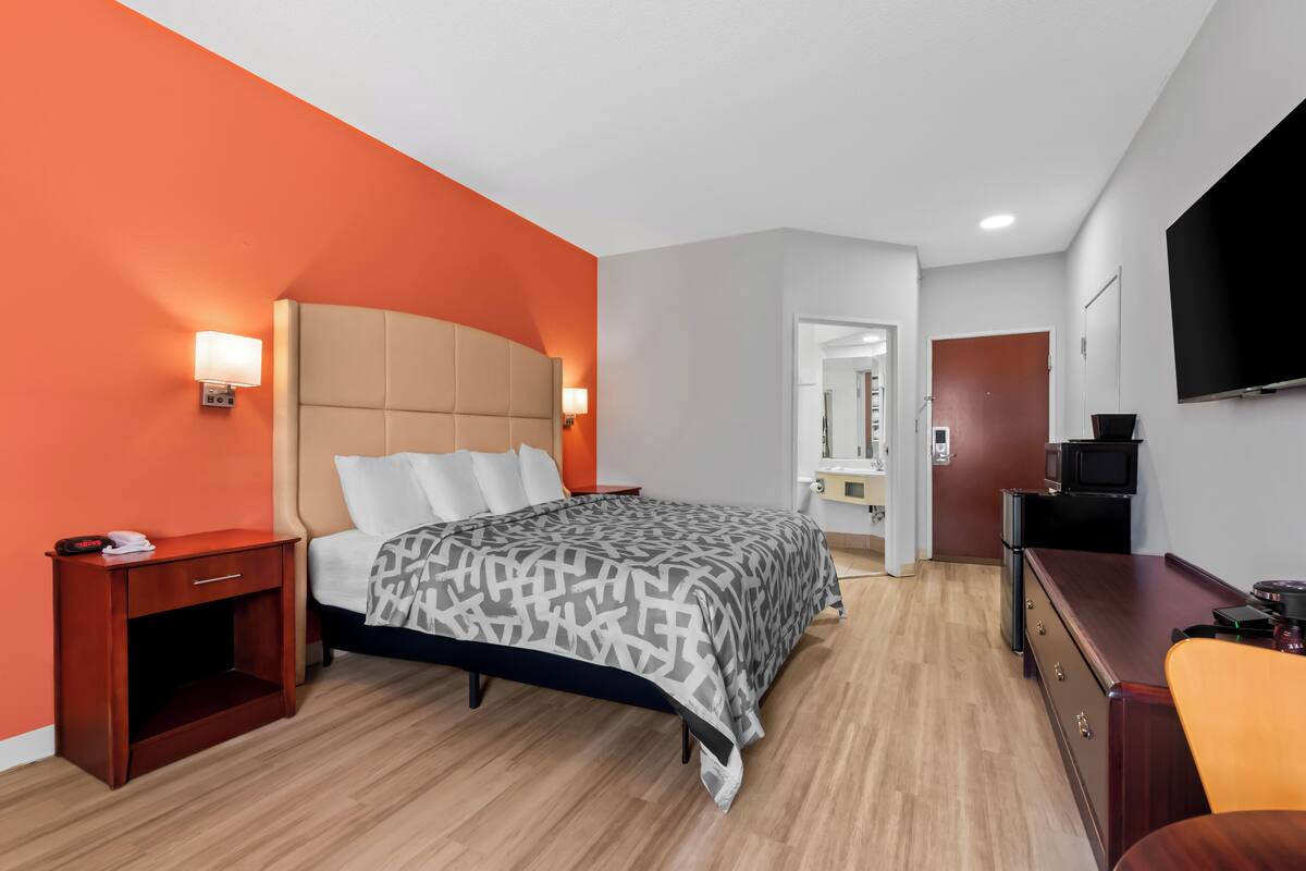 Quarto de hotel com cama, mesa de cabeceira, luminária e televisão.