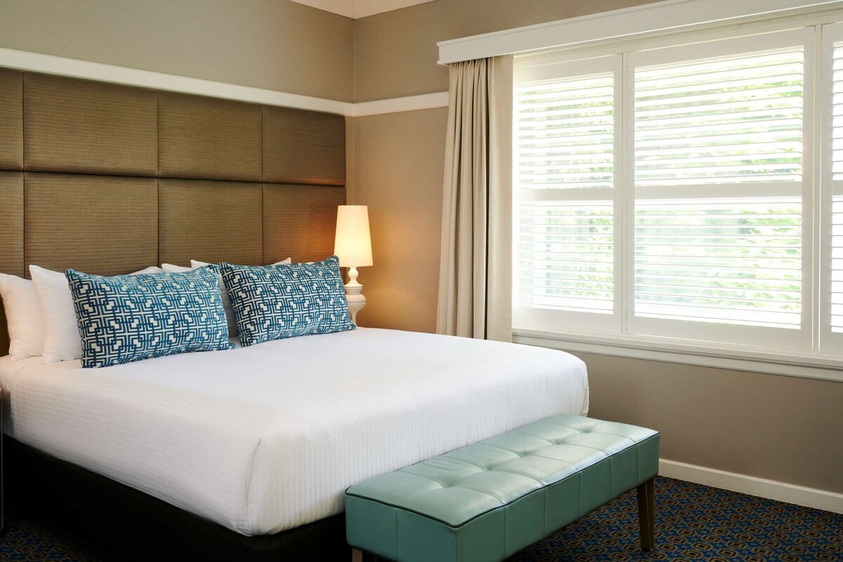 Quarto de hotel com uma cama grande, cabeceira, janela com persianas, abajur e um banco.