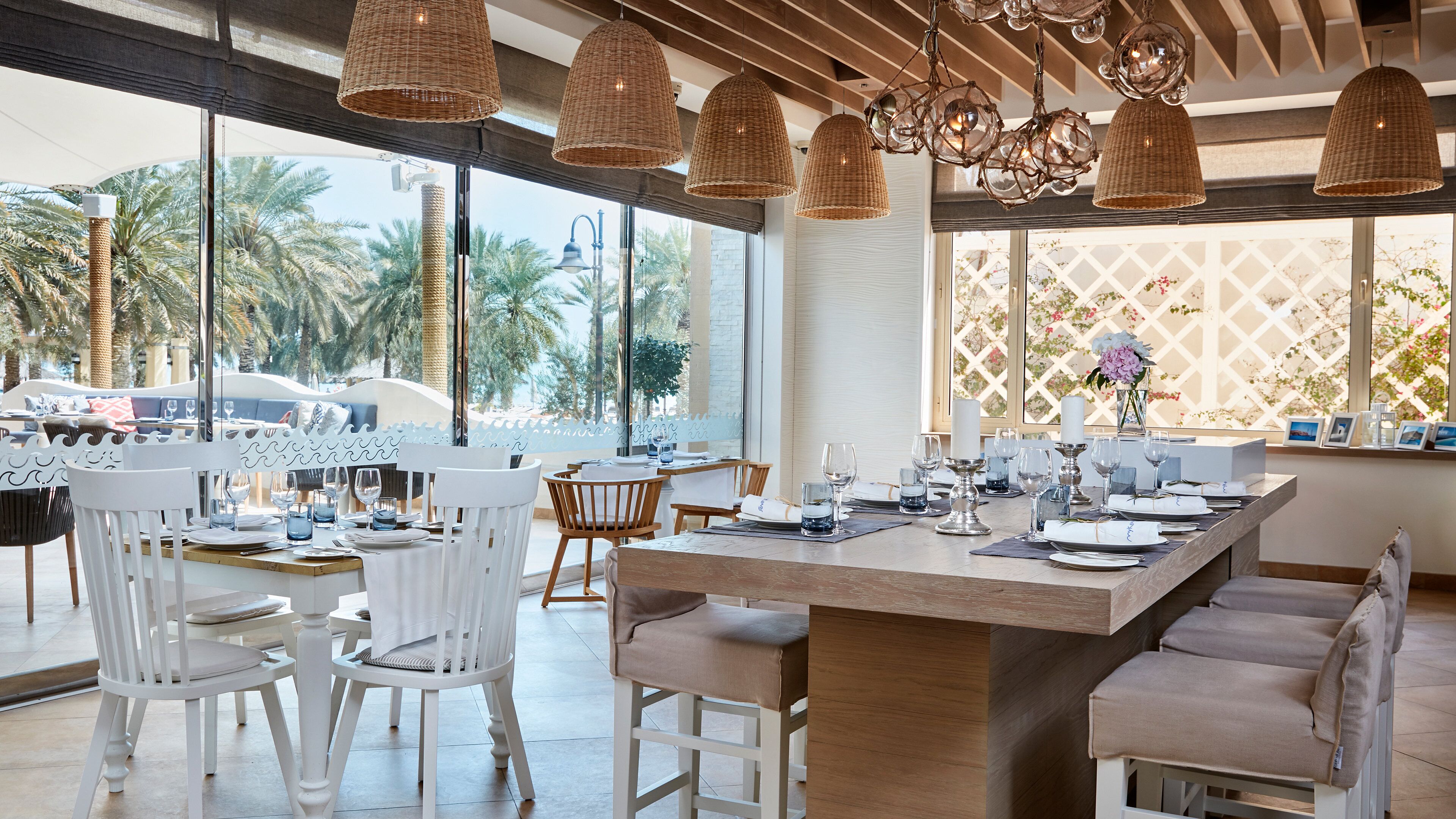Restaurante com uma mesa comprida posta para refeição, cadeiras brancas e luminárias de vime penduradas.