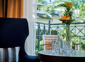 Uma mesa com um vaso de flores e dois copos, uma cadeira, e vista para o verde e edifícios através de uma janela.