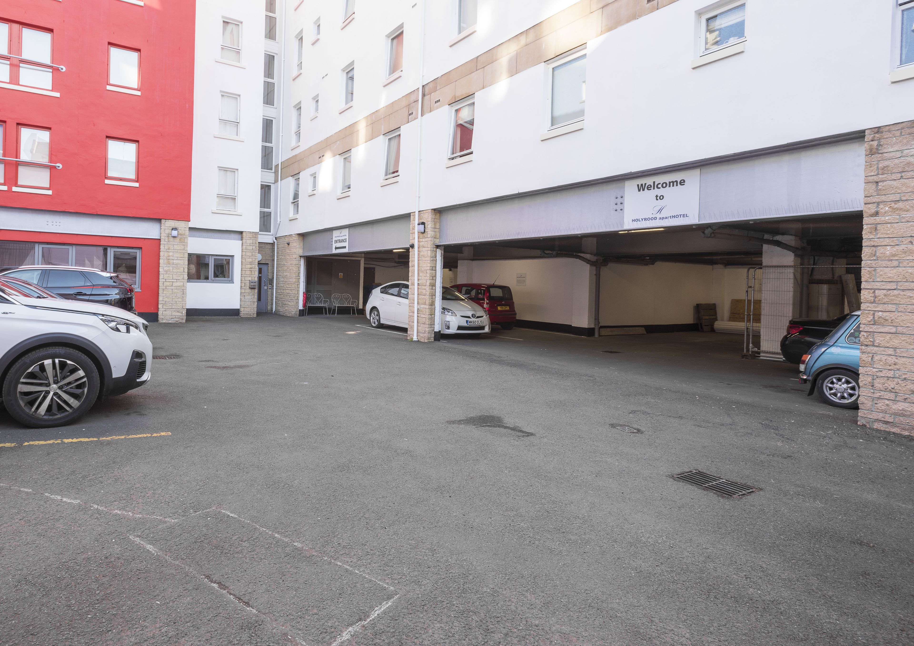 Uma área de estacionamento com vários carros, entrada de um edifício e uma placa de boas-vindas ao apartHOTEL HOLYROOD.
