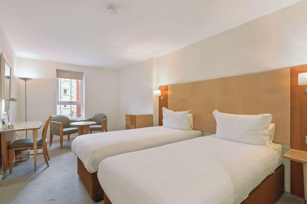 Quarto de hotel com duas camas, uma escrivaninha, uma cadeira e uma janela.
