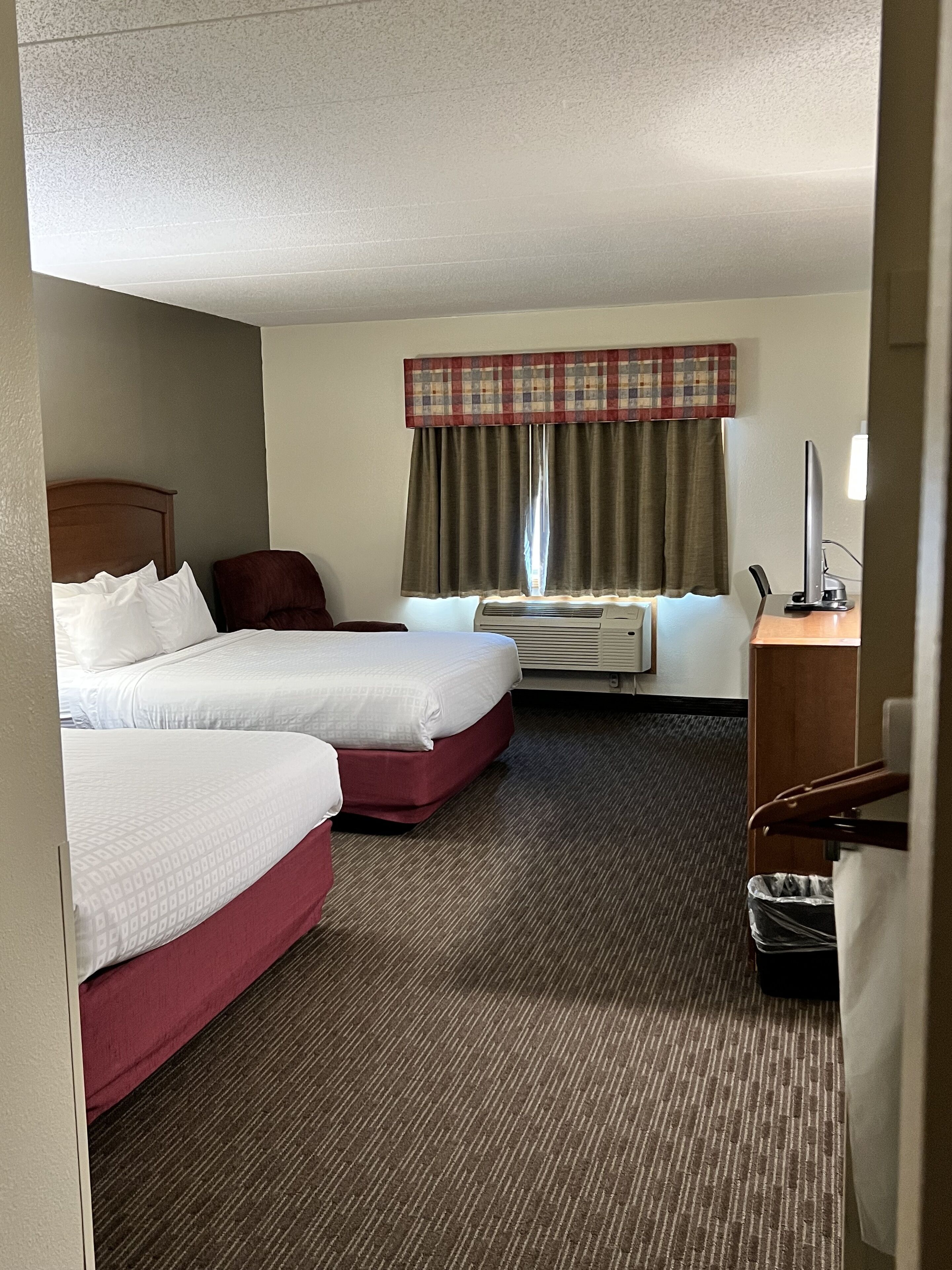 Quarto de hotel com duas camas, uma mesa, uma cadeira, janela com cortinas e ar condicionado fixado na parede.