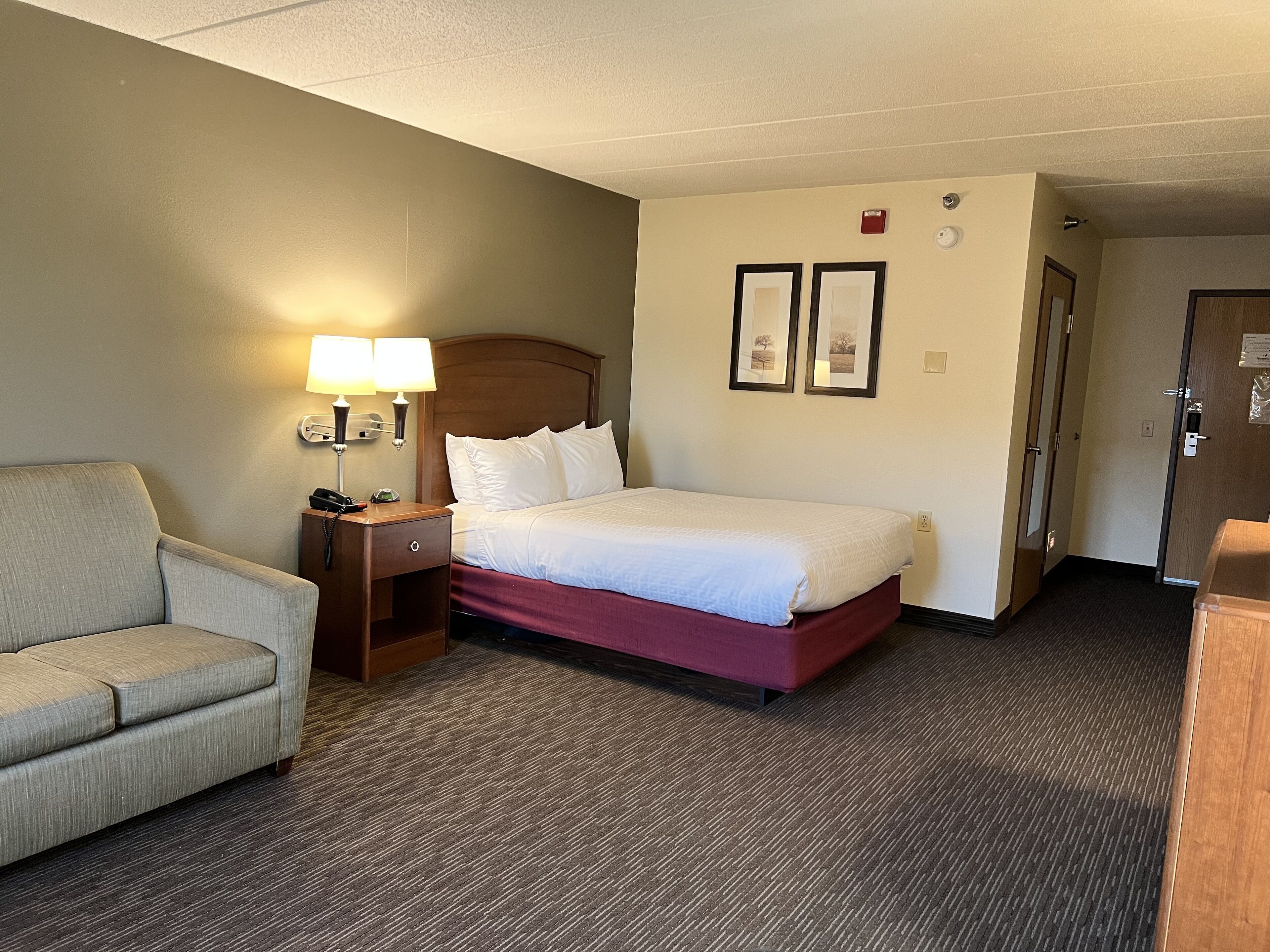 Quarto de hotel com cama, cadeira, mesa de cabeceira com abajur e porta.