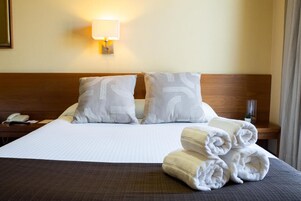 Quarto de hotel com cama bem arrumada, duas almofadas com padrão de folha, luminária de cabeceira e uma pilha de toalhas na cama.