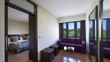 Quarto de hotel moderno com uma cama grande, um sofá roxo, uma mesa pequena e uma janela ampla com vista para o exterior.