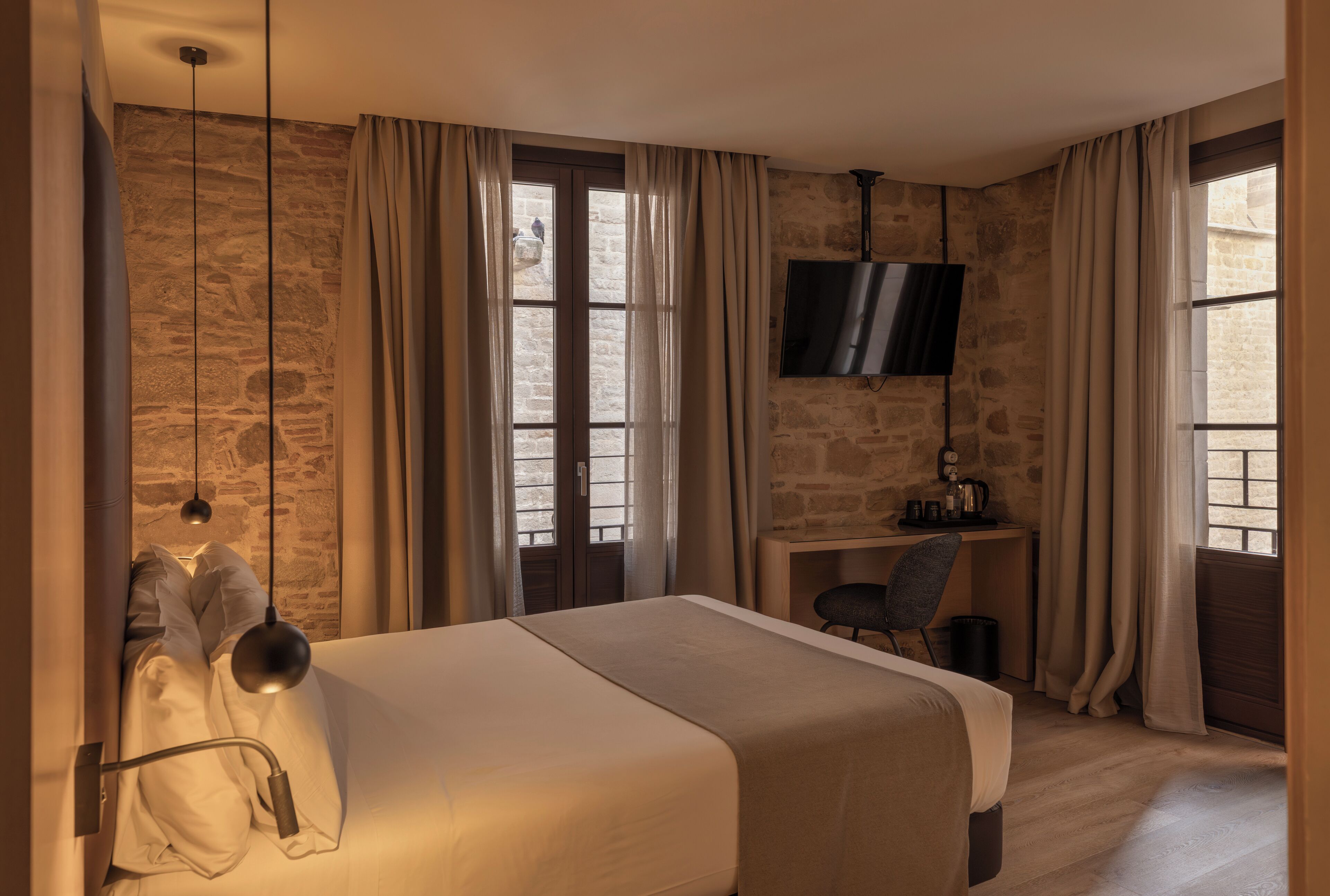 Quarto de hotel com uma cama grande, uma escrivaninha com cadeira, uma televisão e uma parede de pedra.