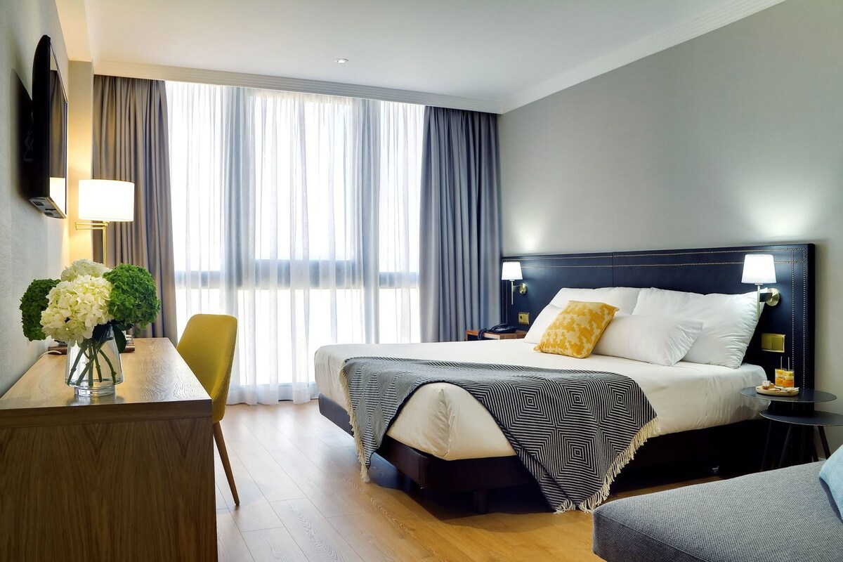 Quarto de hotel com cama, mesas de cabeceira, escrivaninha, cadeira e uma janela grande com cortinas.
